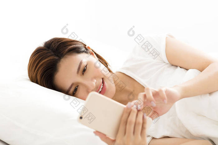 年轻女子看着躺在床上的智能手机 包图企业站