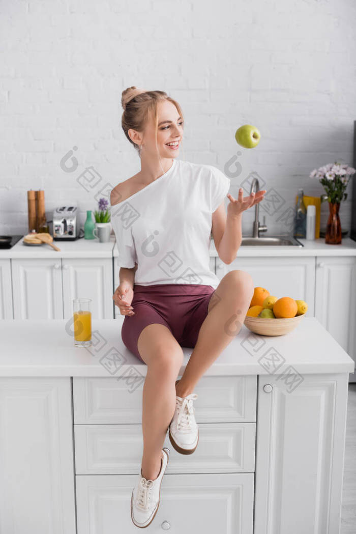快乐的金发女人坐在厨房的桌子上与苹果嬉戏图片 包图网企业站